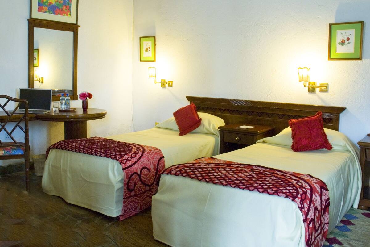 Quarto de hotel com duas camas, uma escrivaninha com um laptop, uma cadeira e uma pequena mesa com um vaso e garrafas de água.