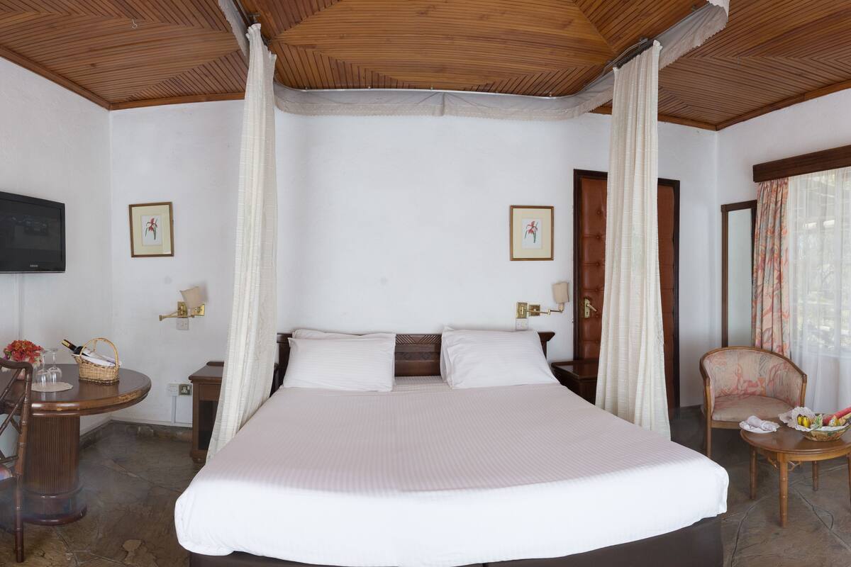 Quarto com uma cama grande, uma cadeira, uma mesinha com uma tigela de frutas e uma televisão na parede.