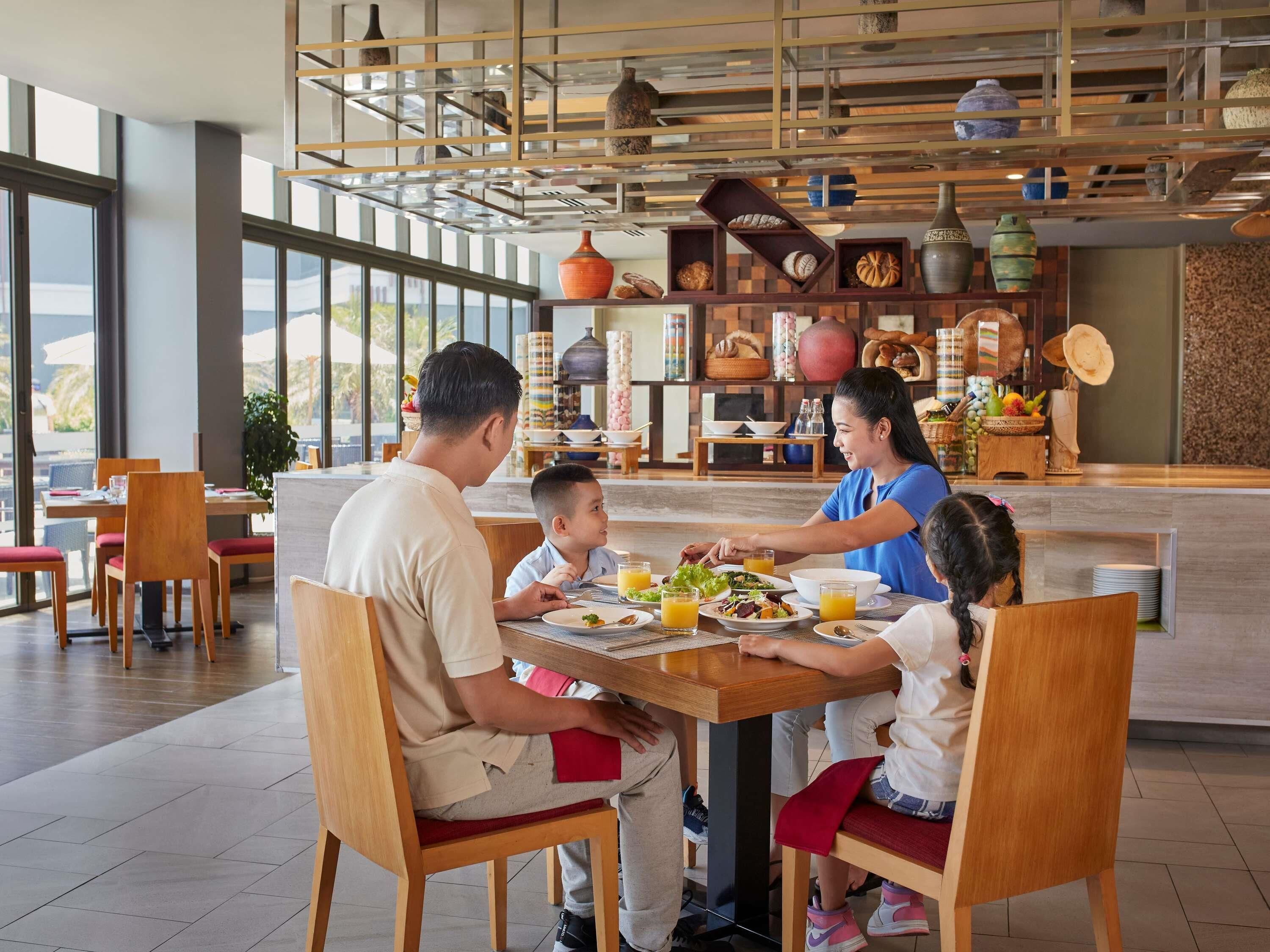 Uma família jantando em um restaurante com decoração moderna, grandes janelas e uma variedade de produtos de panificação à mostra.