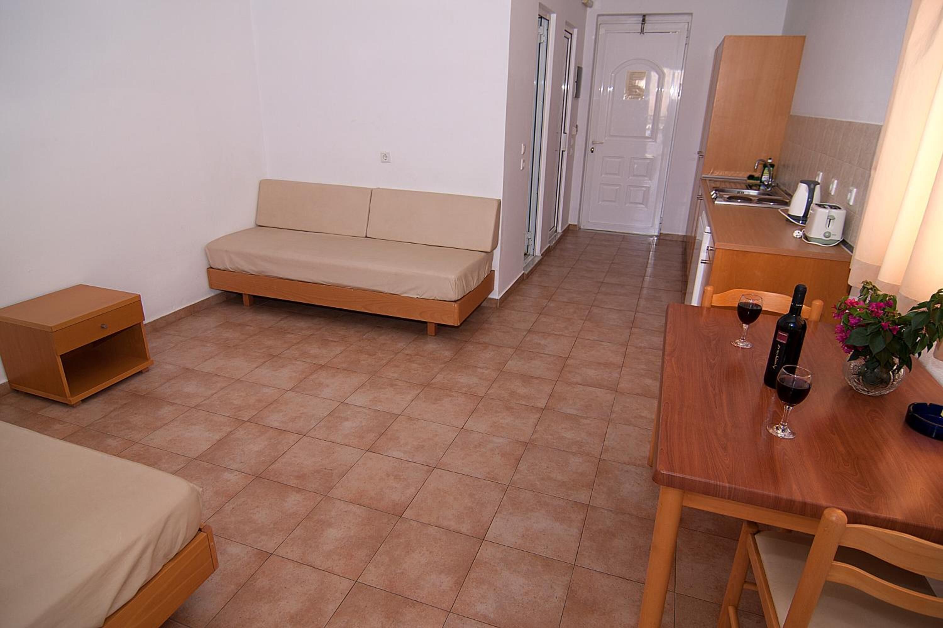 Quarto compacto com sofá, cama, mesa de jantar com cadeiras, pequena cozinha e porta que dá para outro cômodo.