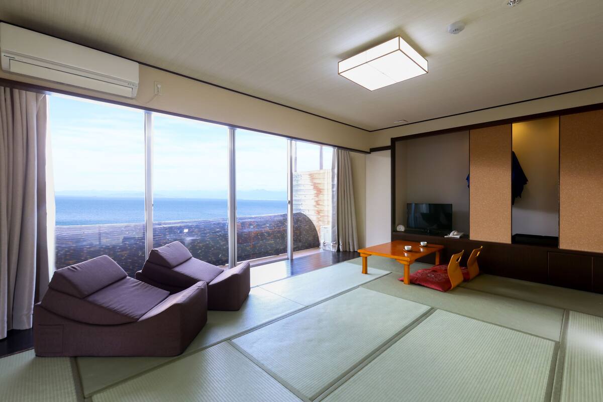 Quarto de hotel moderno com uma grande porta de vidro deslizante para uma varanda, um sofá, uma pequena mesa e uma televisão.