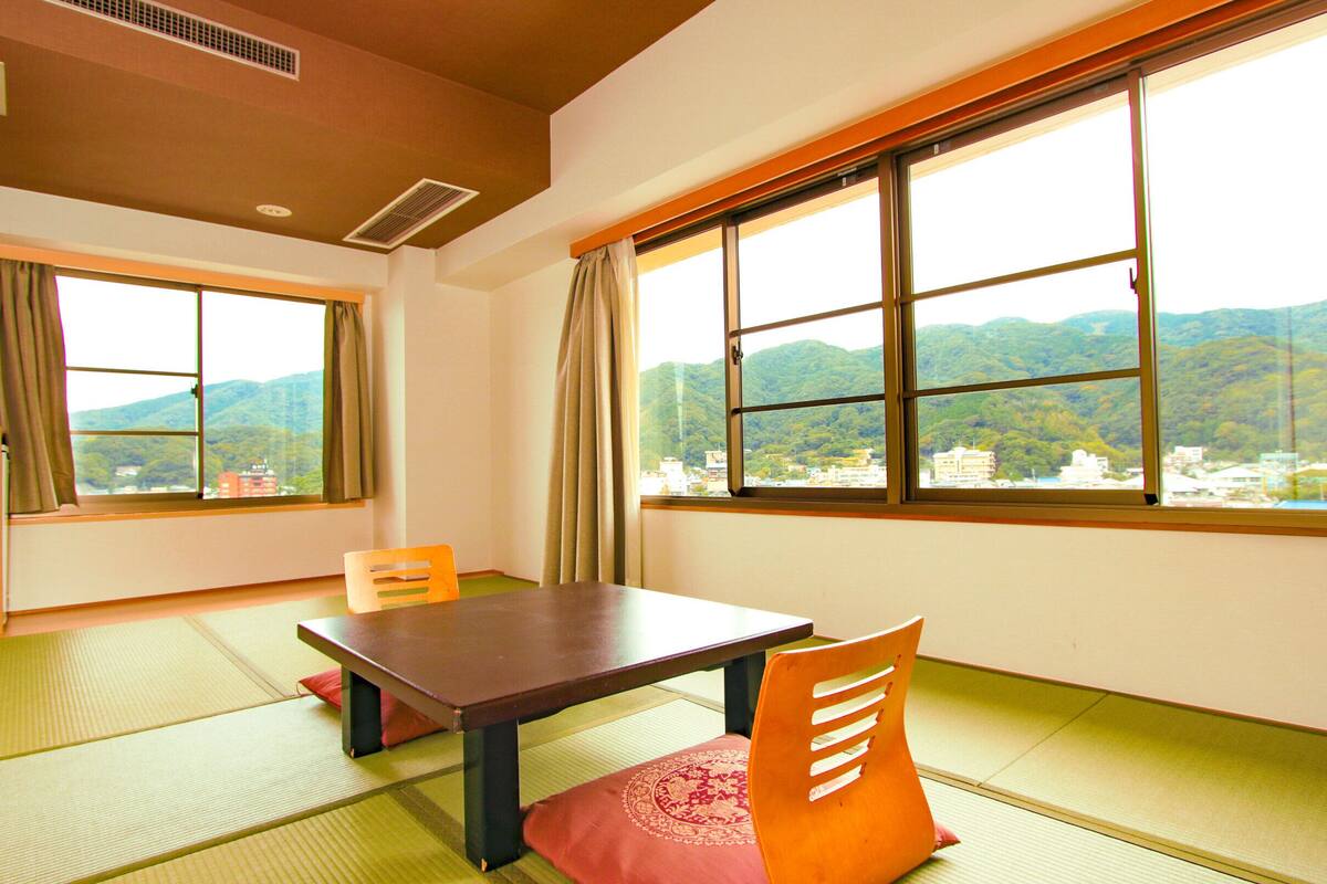 Quarto com esteiras de tatame, uma mesa baixa, uma cadeira e grandes janelas com vista para uma paisagem montanhosa.