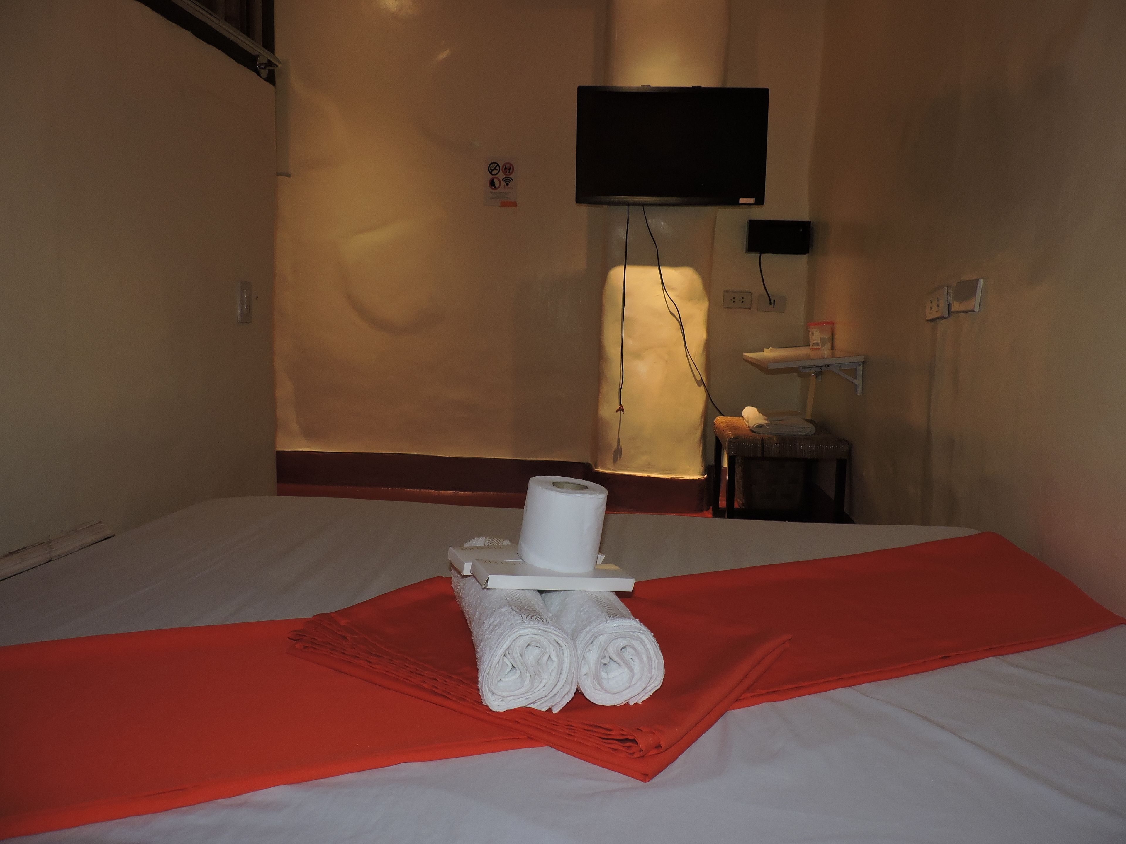 Quarto de hotel com uma cama, televisão na parede e uma mesinha com uma toalha e rolo de papel higiênico.