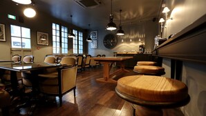 Interior de restaurante com mesas e cadeiras de madeira, iluminação pendente e quadros nas paredes.