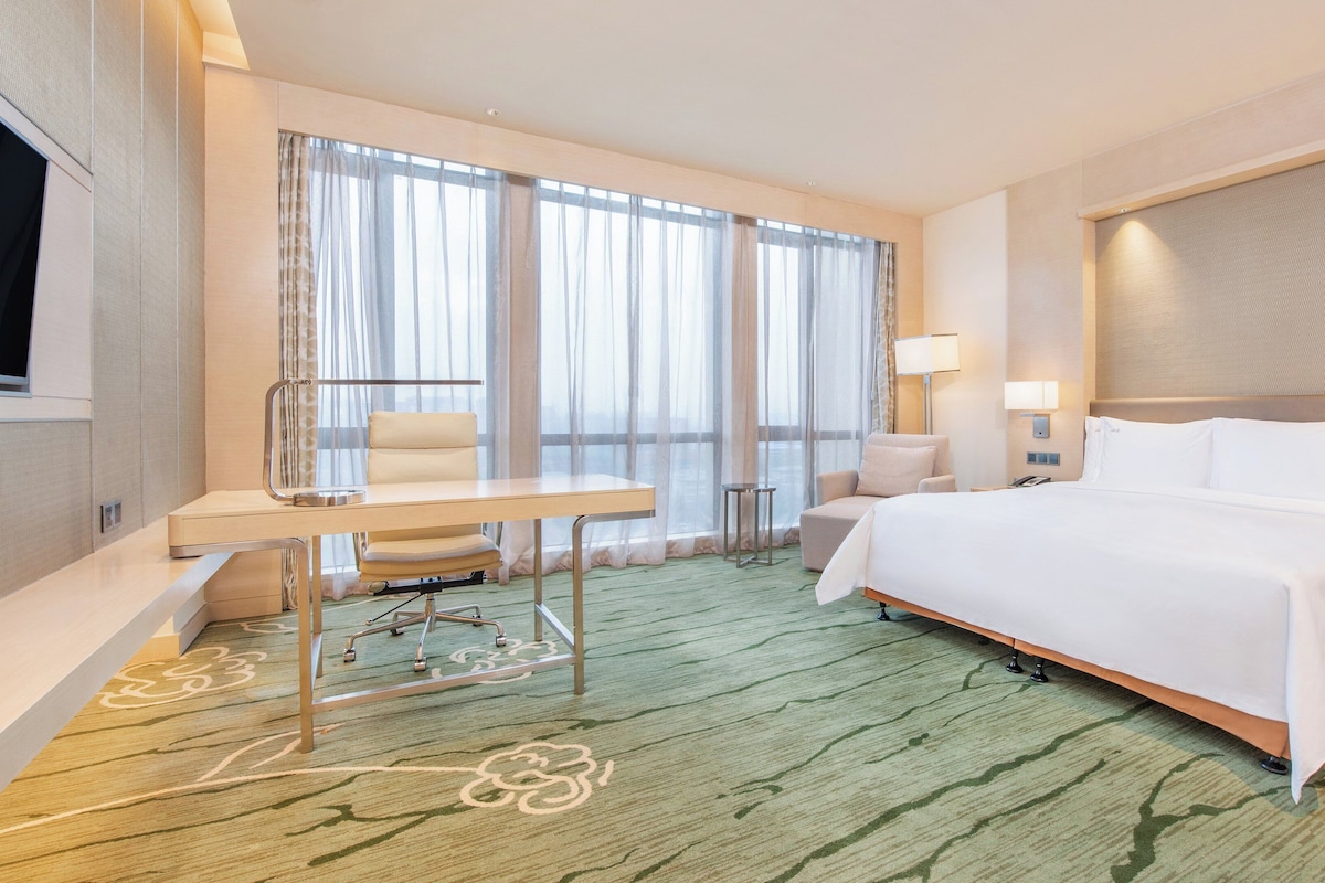 Quarto de hotel com cama, escrivaninha, cadeira e grandes janelas.