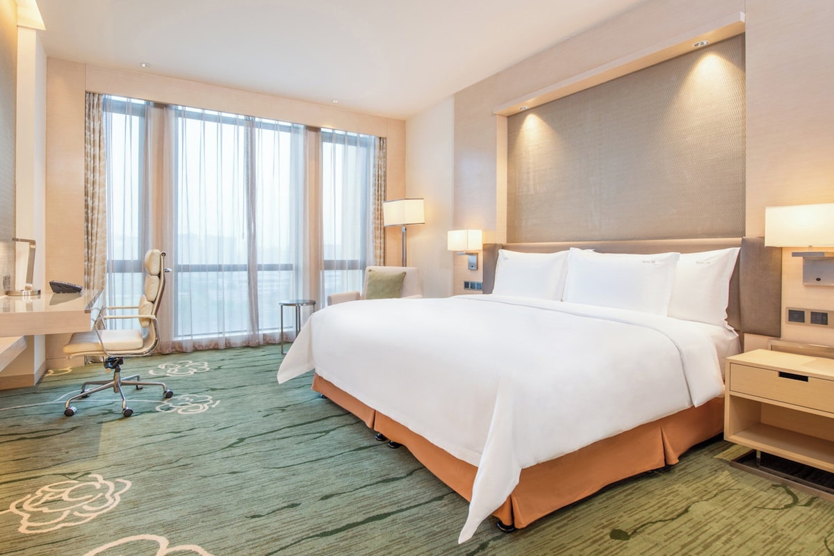 Quarto de hotel com uma cama grande, uma escrivaninha, uma cadeira, uma TV e uma janela com cortinas.