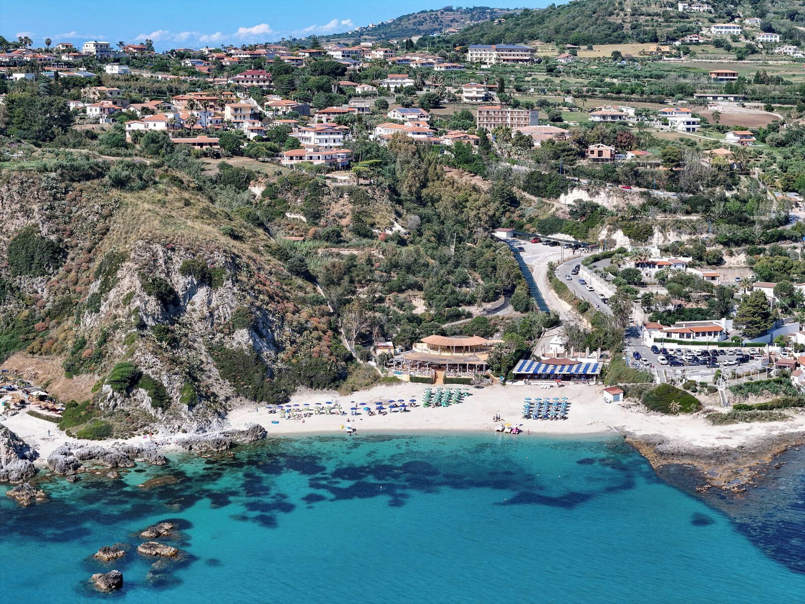Vista aérea de uma vila costeira com praia, casas e uma estrada sinuosa.
