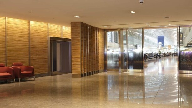 Um lobby moderno com piso reluzente, assentos vermelhos, um elevador de vidro e vista para uma área movimentada.