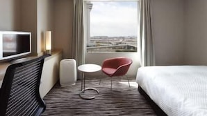 Quarto de hotel com cama, TV, escrivaninha, cadeira e vista da cidade pela janela.