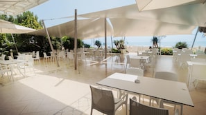 Área de refeições ao ar livre com mesas e cadeiras, piso de azulejos e vista para a praia.