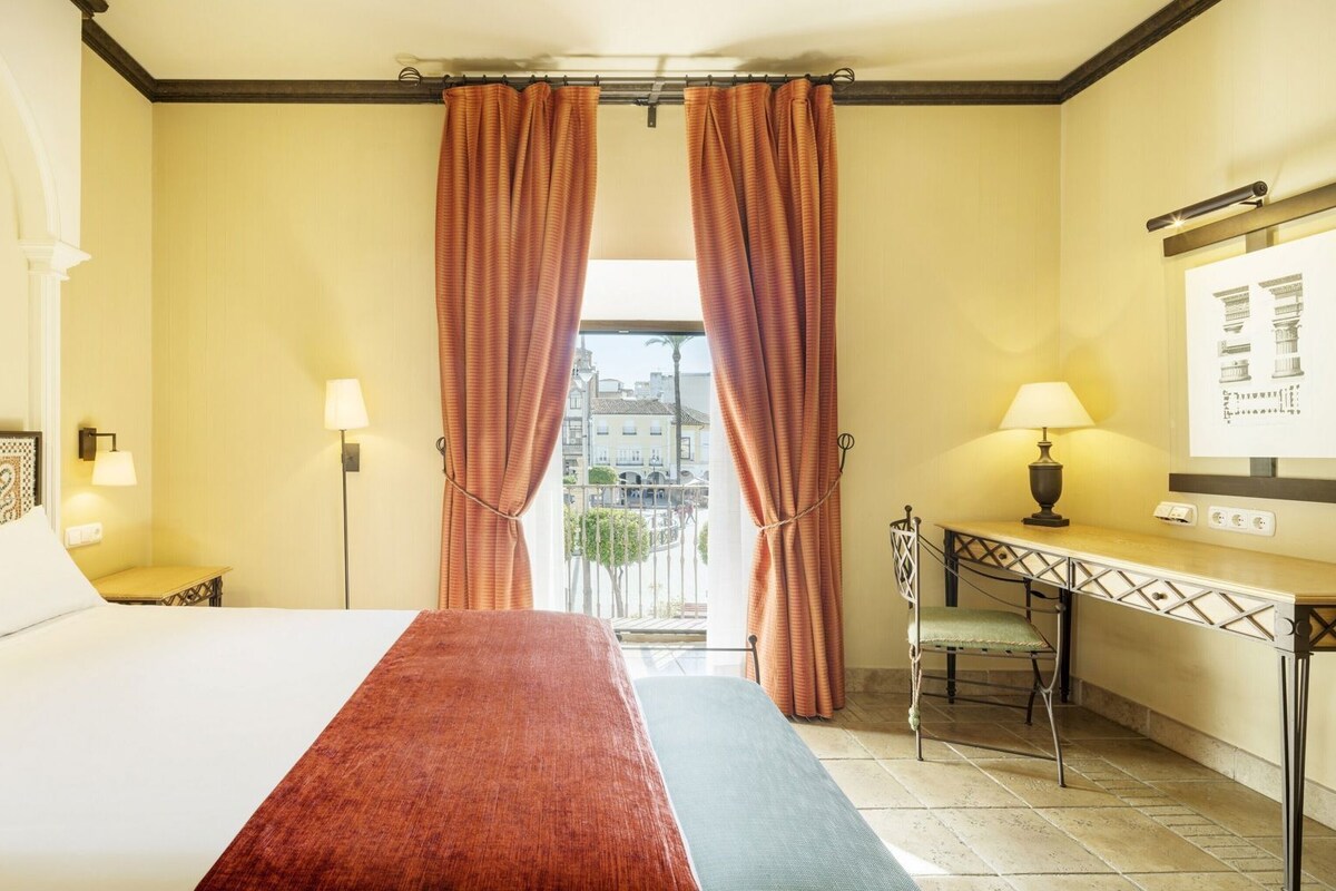 Quarto de hotel com cama, escrivaninha, cadeira, abajur e vista da cidade pela janela.