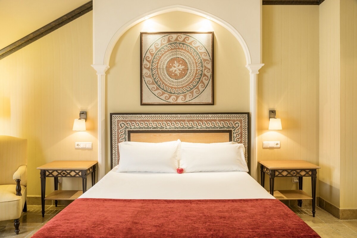 Quarto de hotel com uma cama grande, duas mesas de cabeceira, uma cadeira e um objeto decorativo na parede.
