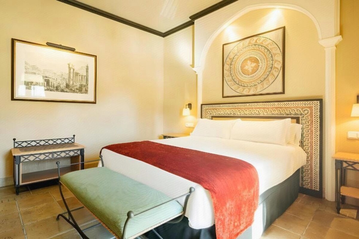 Quarto de hotel com uma cama, um banco, uma mesa de cabeceira e duas luminárias fixadas na parede.