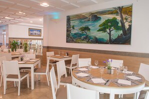 Um restaurante com mesas redondas, cadeiras brancas e uma grande pintura de paisagem na parede.