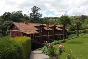 Edifício de tijolos vermelhos com telhado em telhas, rodeado por vegetação exuberante e um gramado bem cuidado.