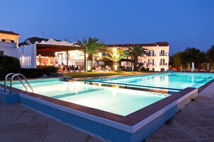 Piscina com deck revestido, cercada por palmeiras e um edifício com varandas múltiplas.