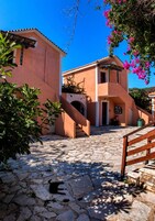 Um pátio pavimentado em pedra com um banco de madeira, um edifício com fachada rosa e um céu azul límpido.