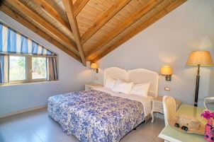 Quarto com teto de madeira, cama com colcha florida, luminárias de cabeceira, uma pequena mesa com abajur e uma janela com cortinas.