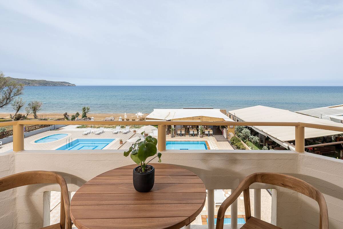 Varanda com mesa e cadeiras de madeira, com vista para uma piscina e praia.