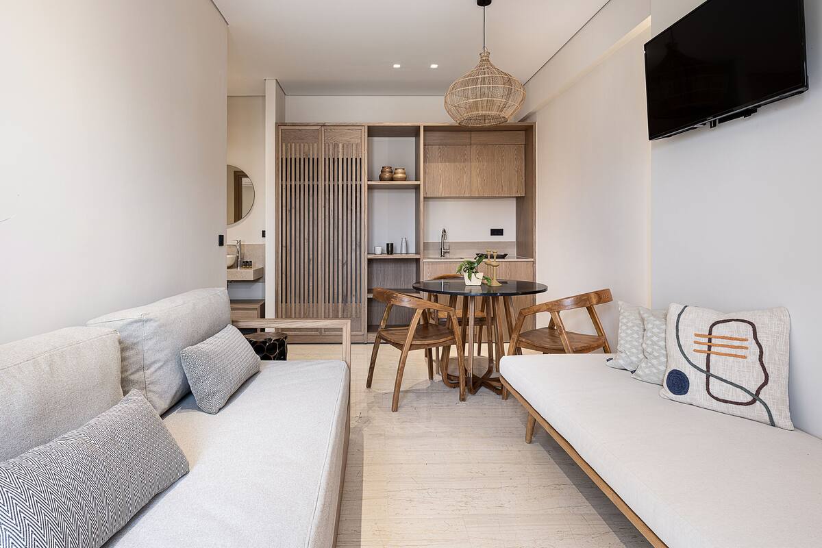 Uma sala de estar moderna com um sofá, uma mesa de jantar com cadeiras, uma TV na parede e uma área de cozinha ao fundo.