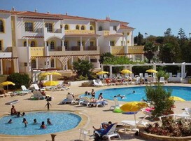 Uma área de piscina com vários espreguiçadeiras e guarda-sóis, rodeada por um edifício com telhado em azulejos e varandas.