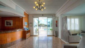 Um lobby de hotel com um balcão de recepção, área de estar com sofá e poltronas, mesa de centro de vidro e vista através das portas de vidro.