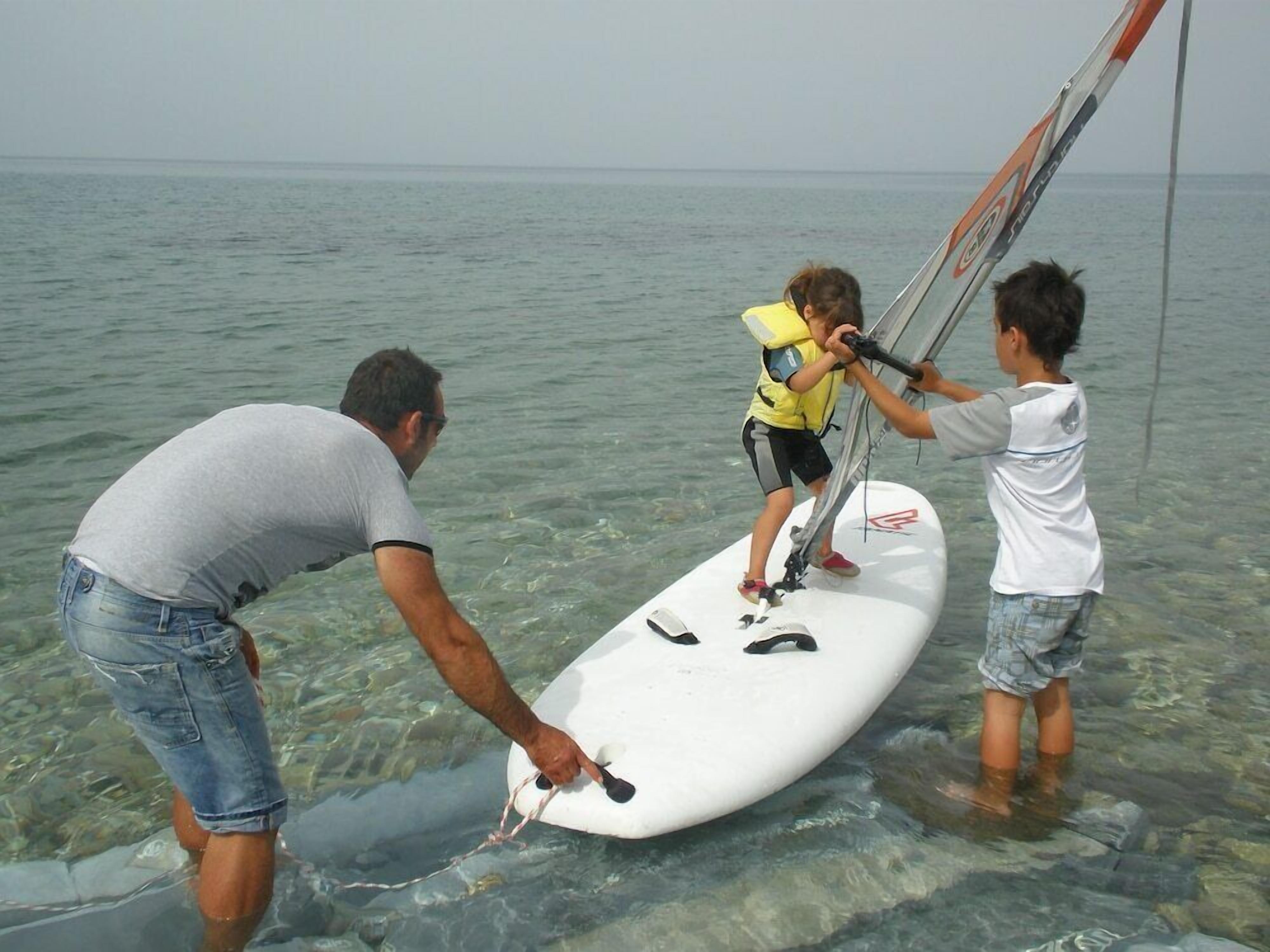 Um homem e duas crianças preparando uma prancha de vela em águas rasas.