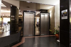 Visão interna de um saguão de hotel com balcão de recepção, dois elevadores e um vaso de planta.