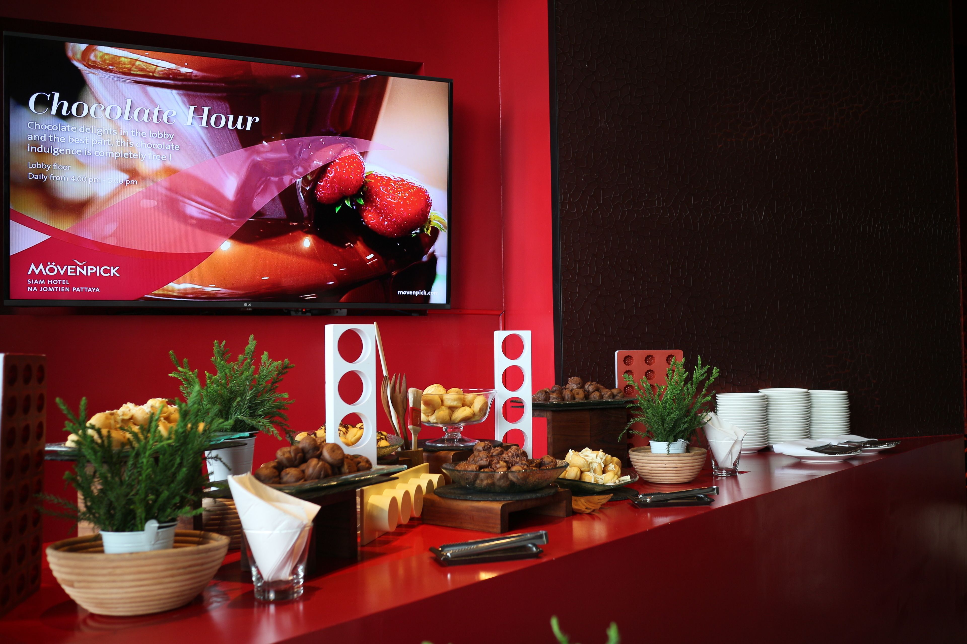Um saguão de hotel com uma televisão exibindo 'Chocolate Hour' e um buffet com diversas opções de comida e decoração.