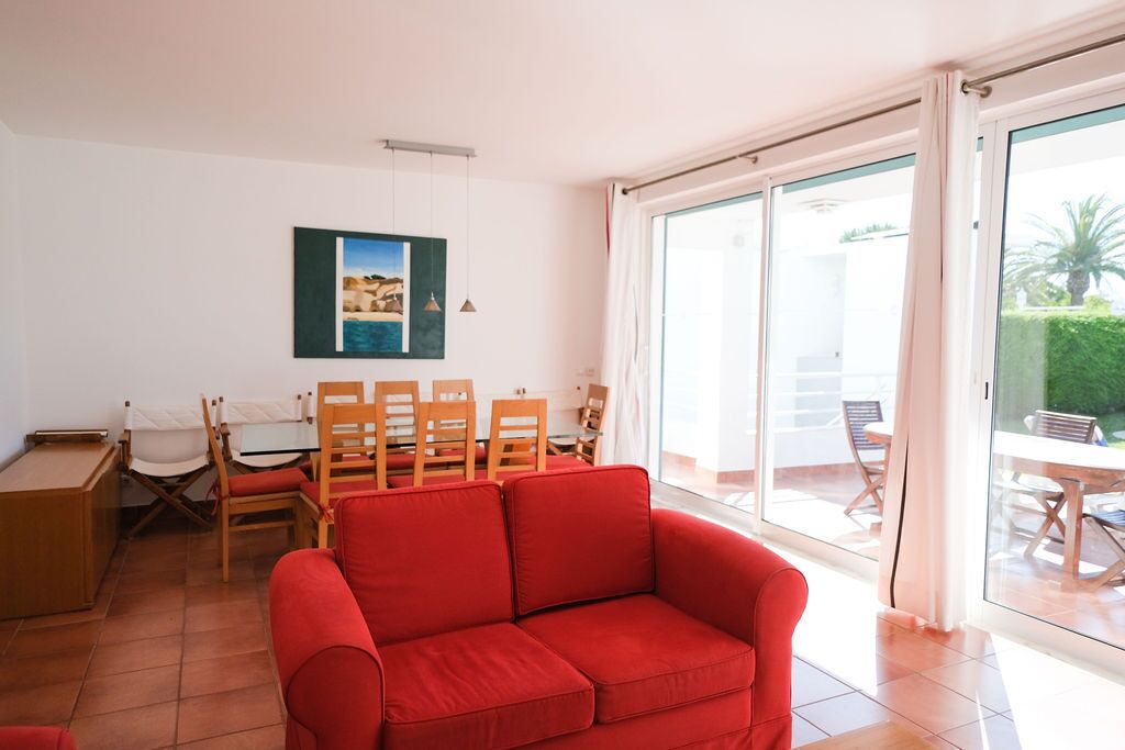 Uma sala de estar iluminada, com um sofá vermelho, cadeiras de madeira, uma mesa de jantar e uma grande porta de vidro deslizante que dá para um pátio externo.