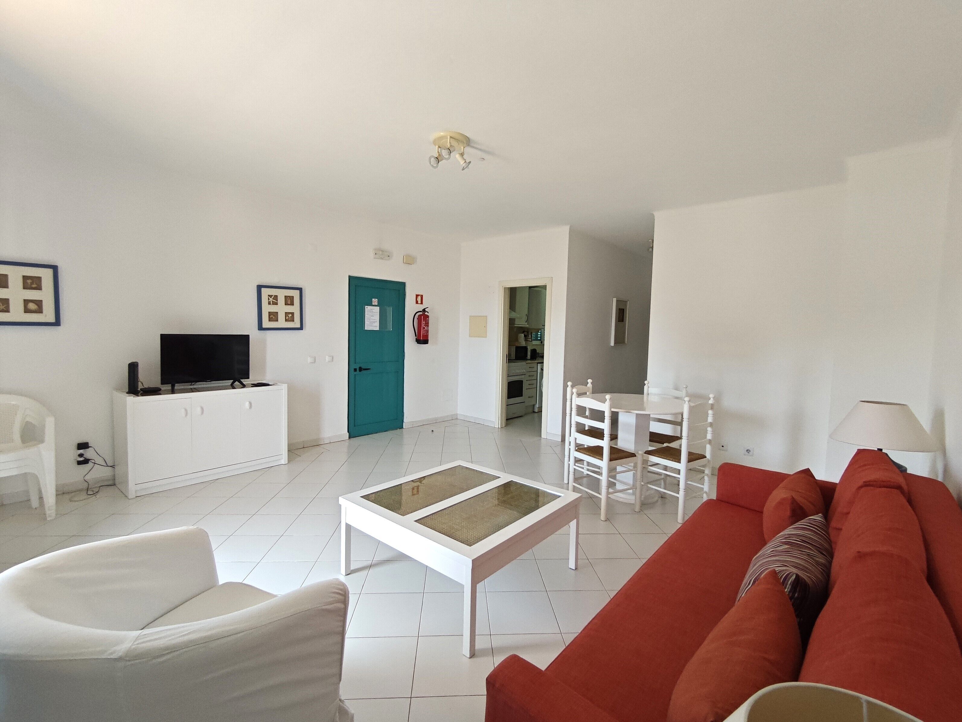 Sala de estar com um sofá branco, um sofá vermelho, uma mesa de centro branca, uma televisão sobre um móvel branco e uma porta verde-água.