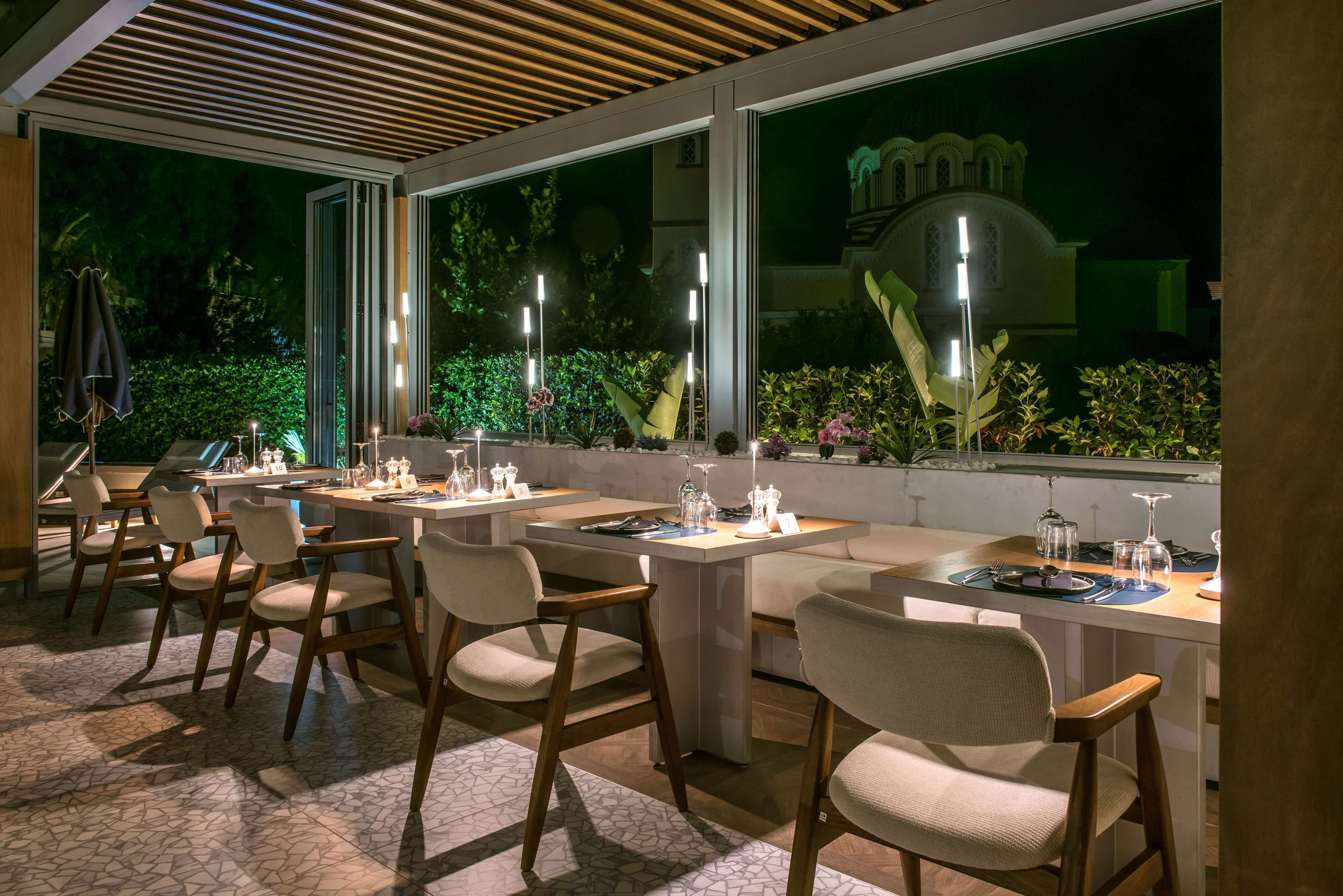 Uma área de refeições ao ar livre com mesas e cadeiras, iluminada por luzes verticais, e grandes janelas com vista para vegetação e um edifício.