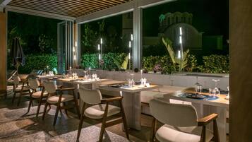 Uma área de refeições ao ar livre com mesas e cadeiras, iluminada por luzes verticais, e grandes janelas com vista para vegetação e um edifício.