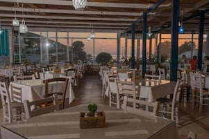 Um restaurante amplo com várias mesas e cadeiras, janelas grandes que oferecem vista para o pôr do sol e luminárias pendentes.