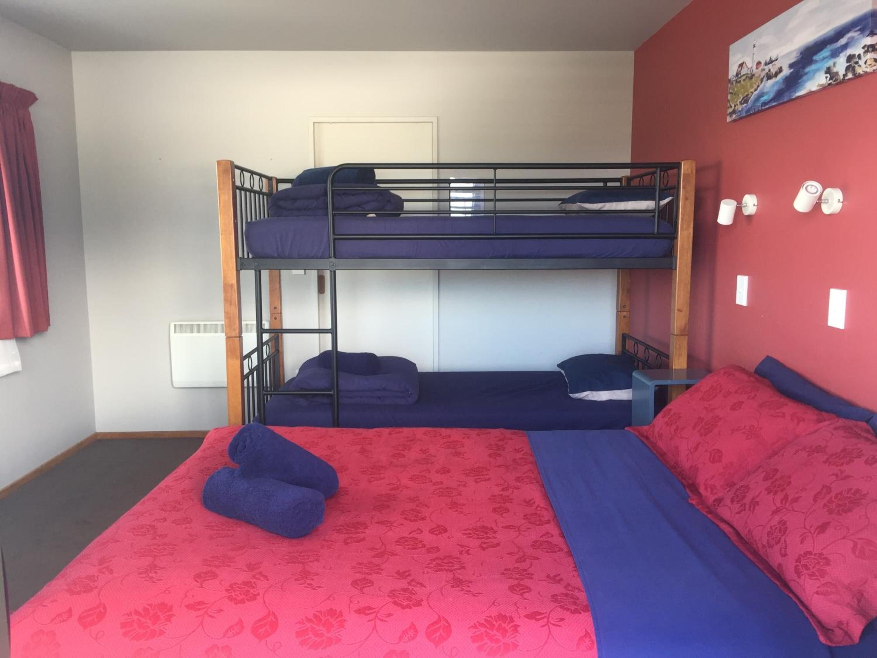 Quarto com beliche, colcha vermelha e azul, ar condicionado branco e um quadro na parede.