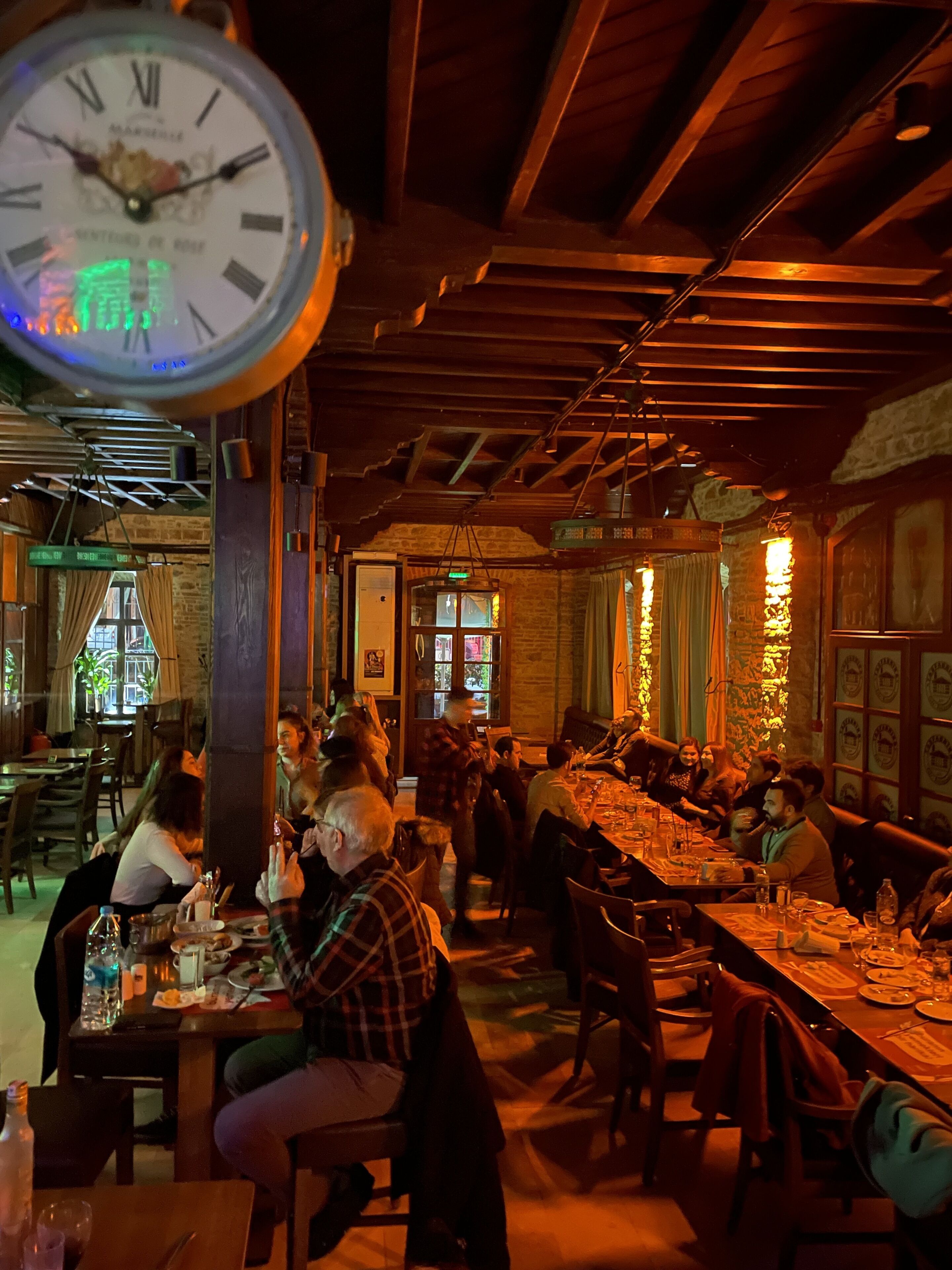 Restaurante com vigas de madeira, paredes de pedra e um relógio grande. Mesas montadas com pratos, copos e garrafas.