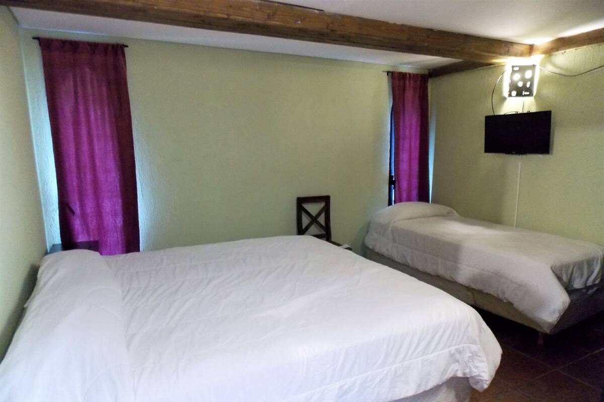 Quarto com duas camas de solteiro, televisão fixada na parede e uma viga de madeira no teto.