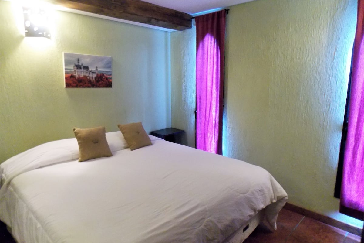 Cama bem arrumada com duas almofadas, uma mesa de cabeceira pequena, uma luminária na parede e um quadro.