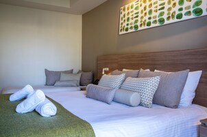 Quarto de hotel com duas camas, roupa de cama cinza, almofadas estampadas e um cobertor verde.