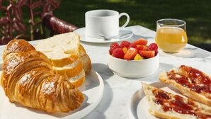 Uma mesa com croissants, morangos, uma xícara e um copo de suco, com um edifício ao fundo.