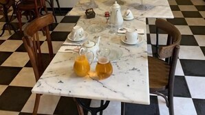 Uma sala de jantar com piso xadrez, uma mesa de mármore posta com serviço de chá, frutas e doces.