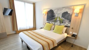 Quarto de hotel com cama, televisão, quadro e secretária com abajur.