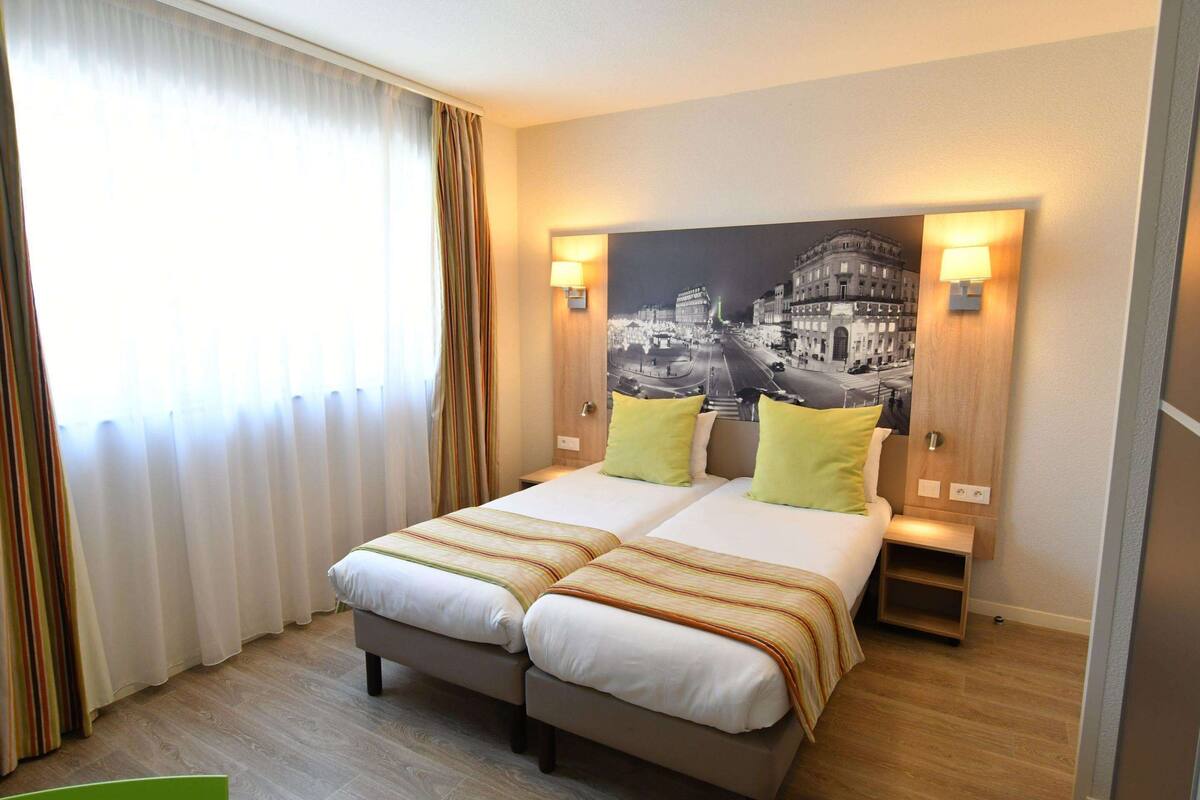 Quarto de hotel com duas camas, uma grande pintura de paisagem urbana na parede e duas luminárias de cabeceira.