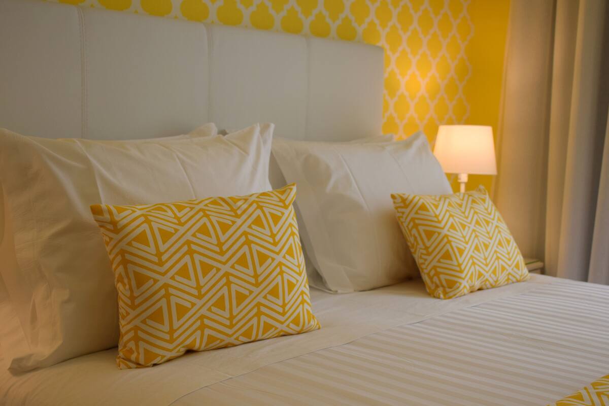 Quarto de hotel com uma cama, duas almofadas estampadas, cabeceira e mesa de cabeceira com abajur.