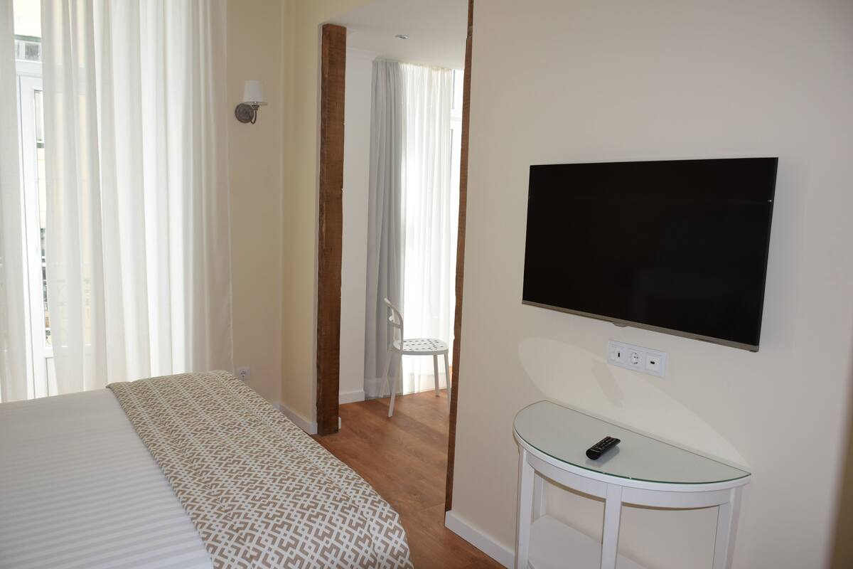 Quarto de hotel com cama, televisão na parede, uma mesinha e uma cadeira.