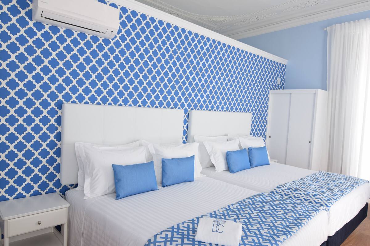 Quarto de hotel com duas camas, cabeceiras brancas e papel de parede estampado em azul.