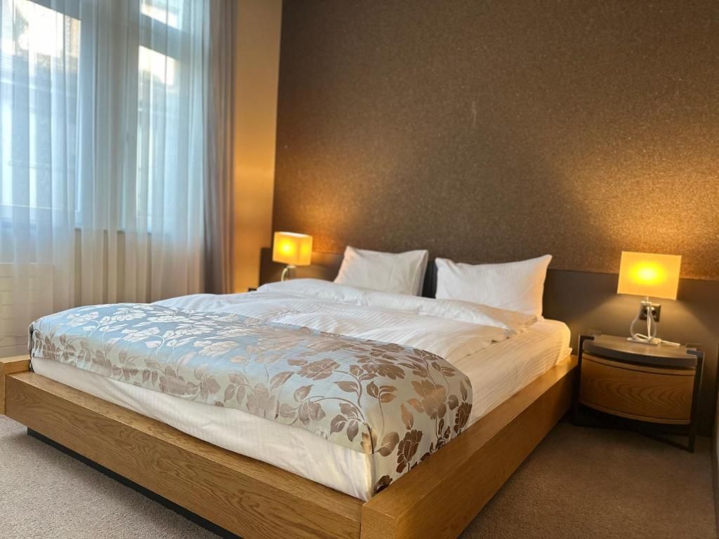 Quarto de hotel com uma cama grande, duas luminárias de cabeceira e vista para o exterior através de cortinas translúcidas.