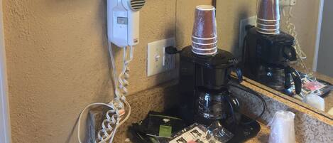 Casa de banho de hotel com lavatório, espelho, porta-toalhas com toalhas dobradas, secador de cabelo, máquina de café e um tabuleiro com diversos objetos.