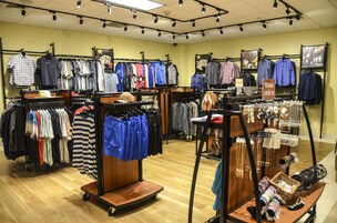 Uma loja de roupas com várias araras de camisas e calças, um expositor de joias e um aviso de liquidação.