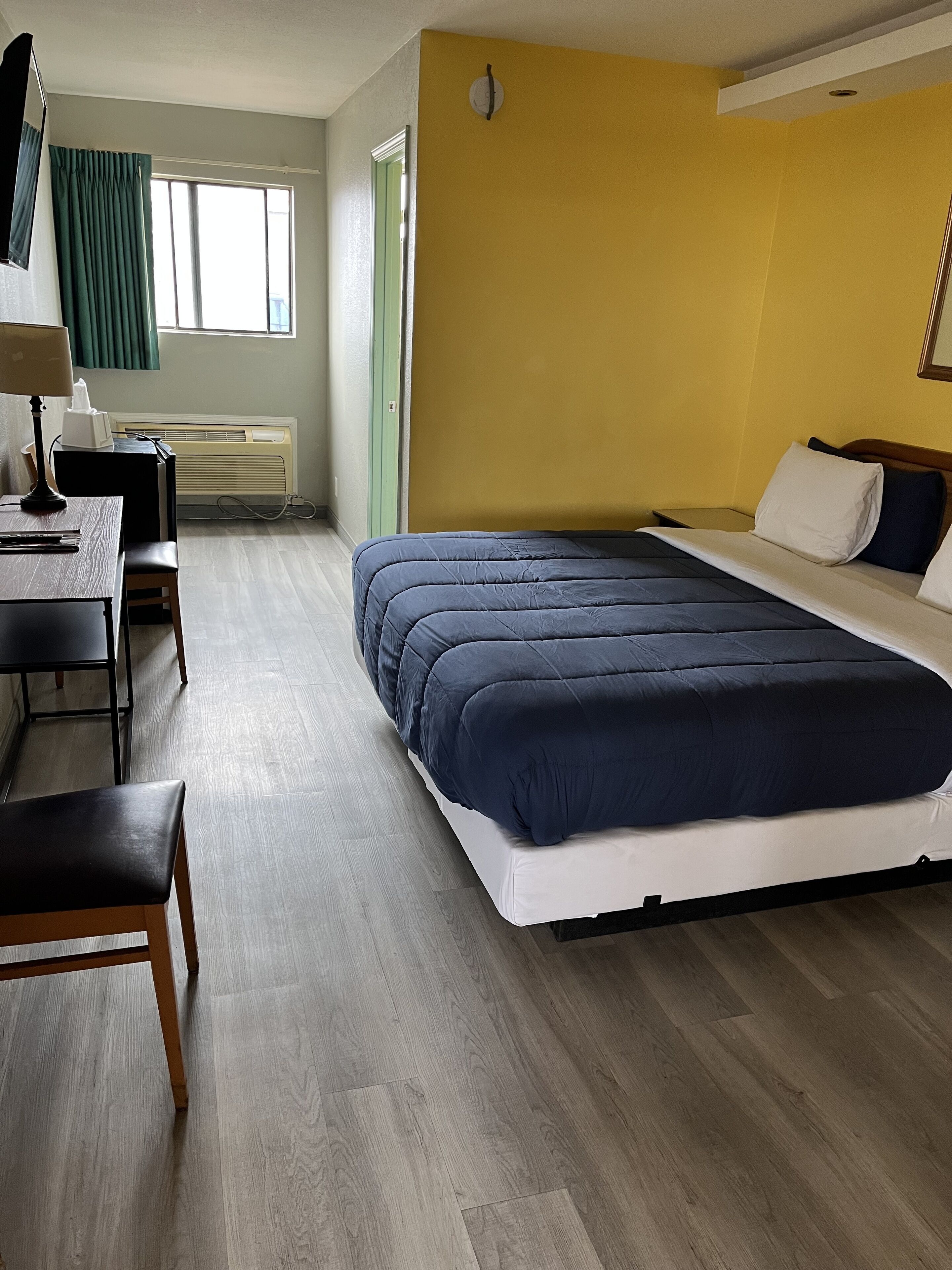 Quarto de hotel com cama, escrivaninha, cadeira, luminária e janela com cortinas.